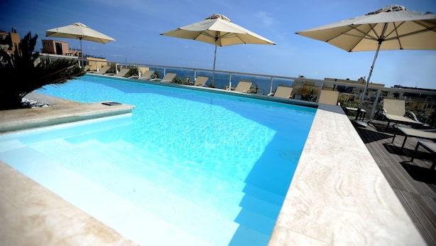 Piscina com espreguiçadeiras e guarda-sóis, cercada por edifícios.