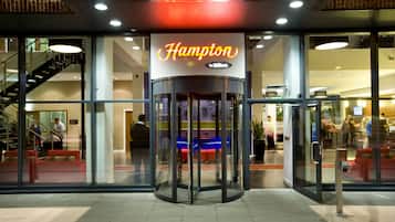 Entrada de um hotel com porta giratória, portas de vidro e uma placa com a inscrição 'Hampton by Hilton'.