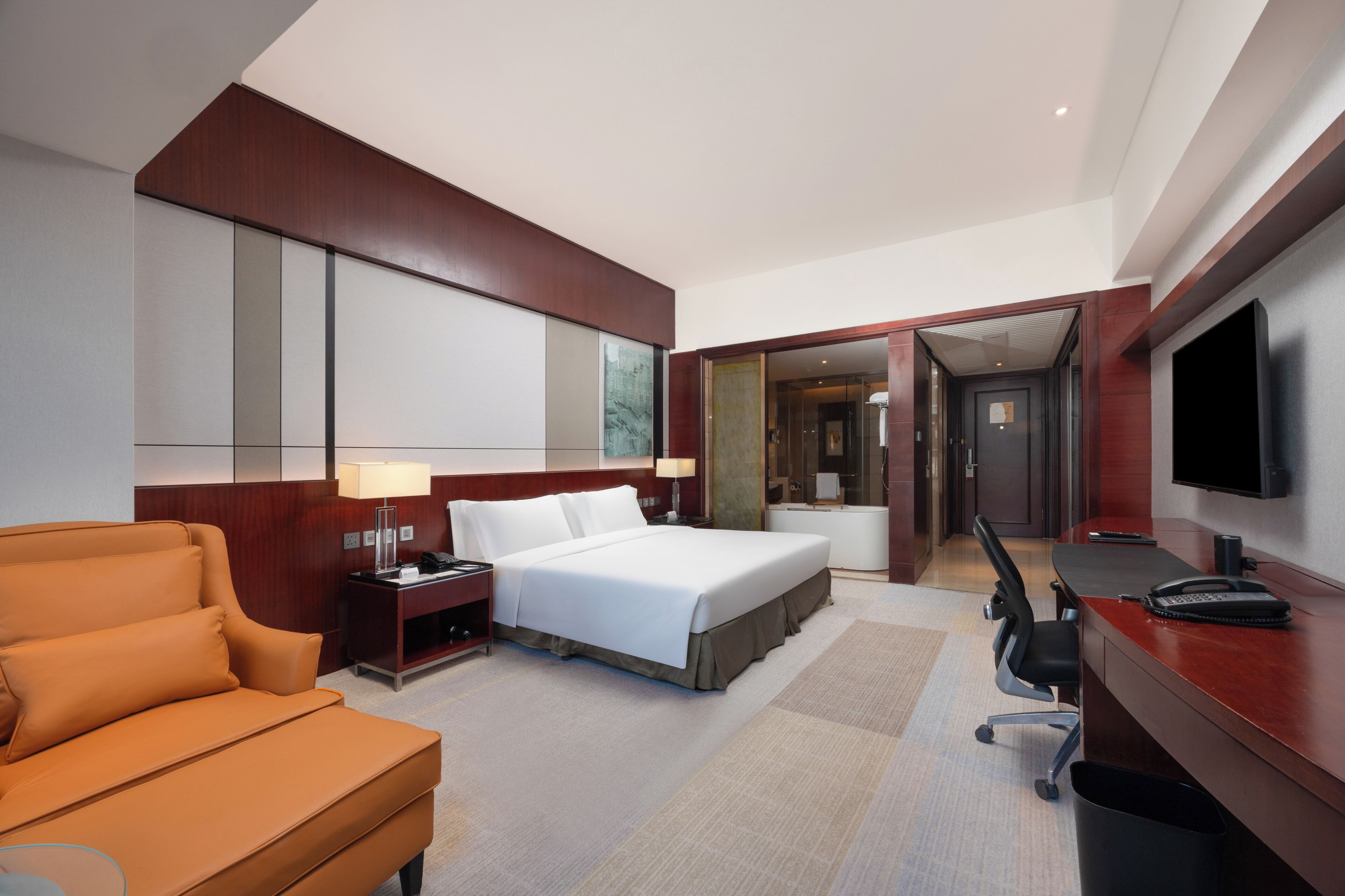 Quarto de hotel com cama, escrivaninha, televisão, sofá e banheiro visível por uma porta aberta.