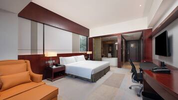 Quarto de hotel com cama, escrivaninha, televisão, sofá e banheiro visível por uma porta aberta.
