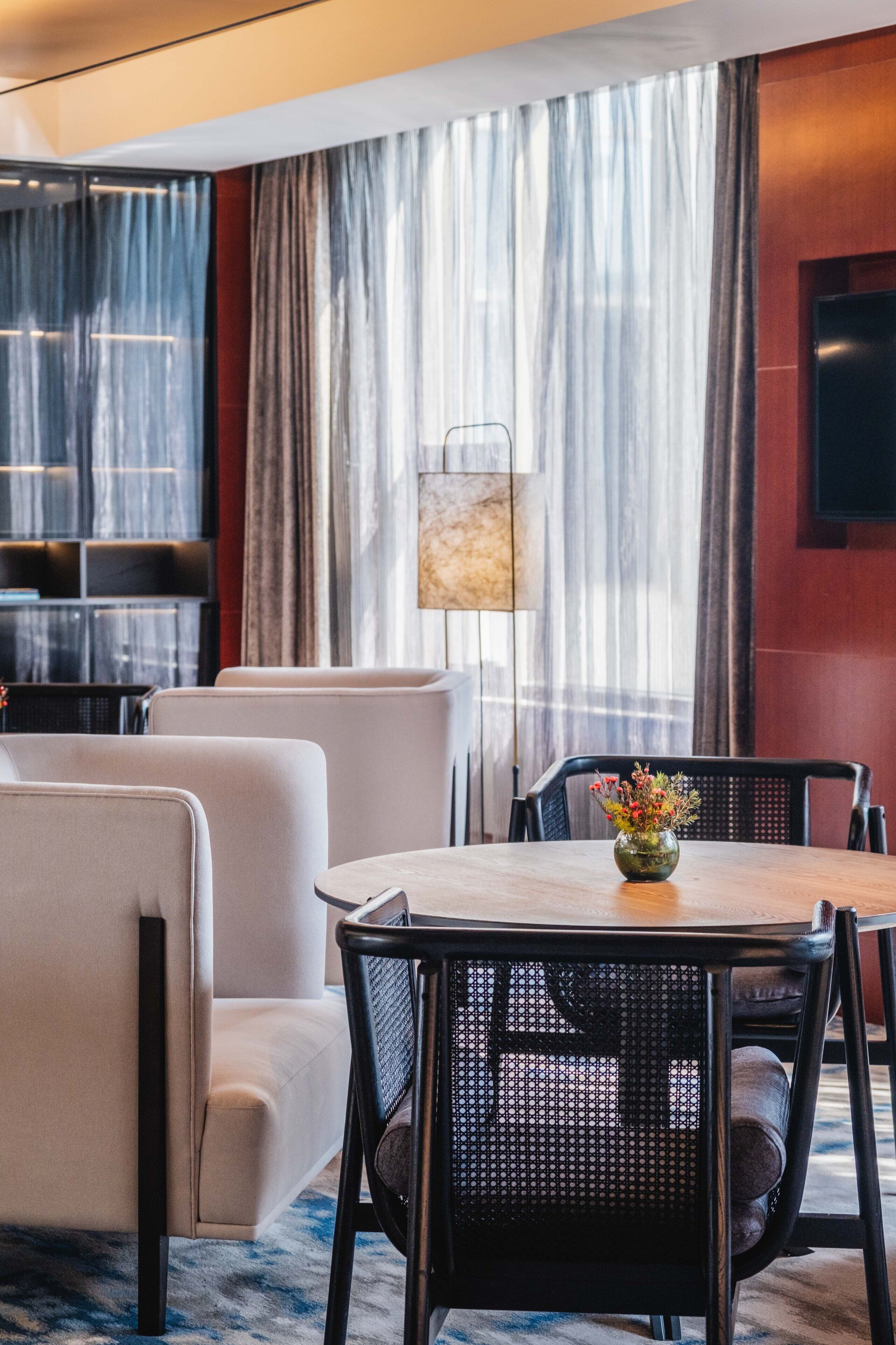 Quarto de hotel com área de jantar, com mesa redonda, cadeiras, um vaso de flores e um abajur.