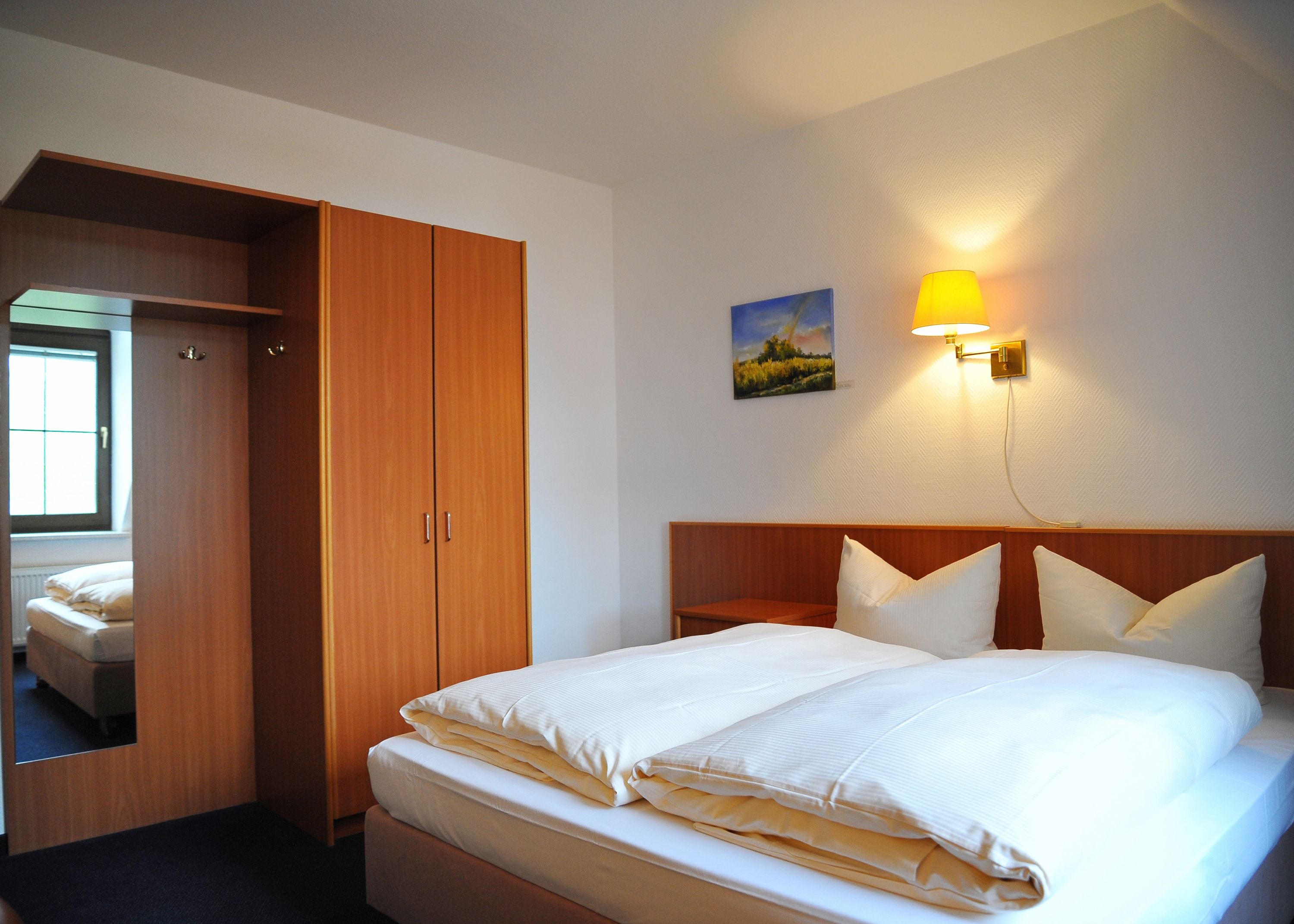 Quarto de hotel com cama, guarda-roupa de madeira, mesa de cabeceira, luminária na parede e uma janela.