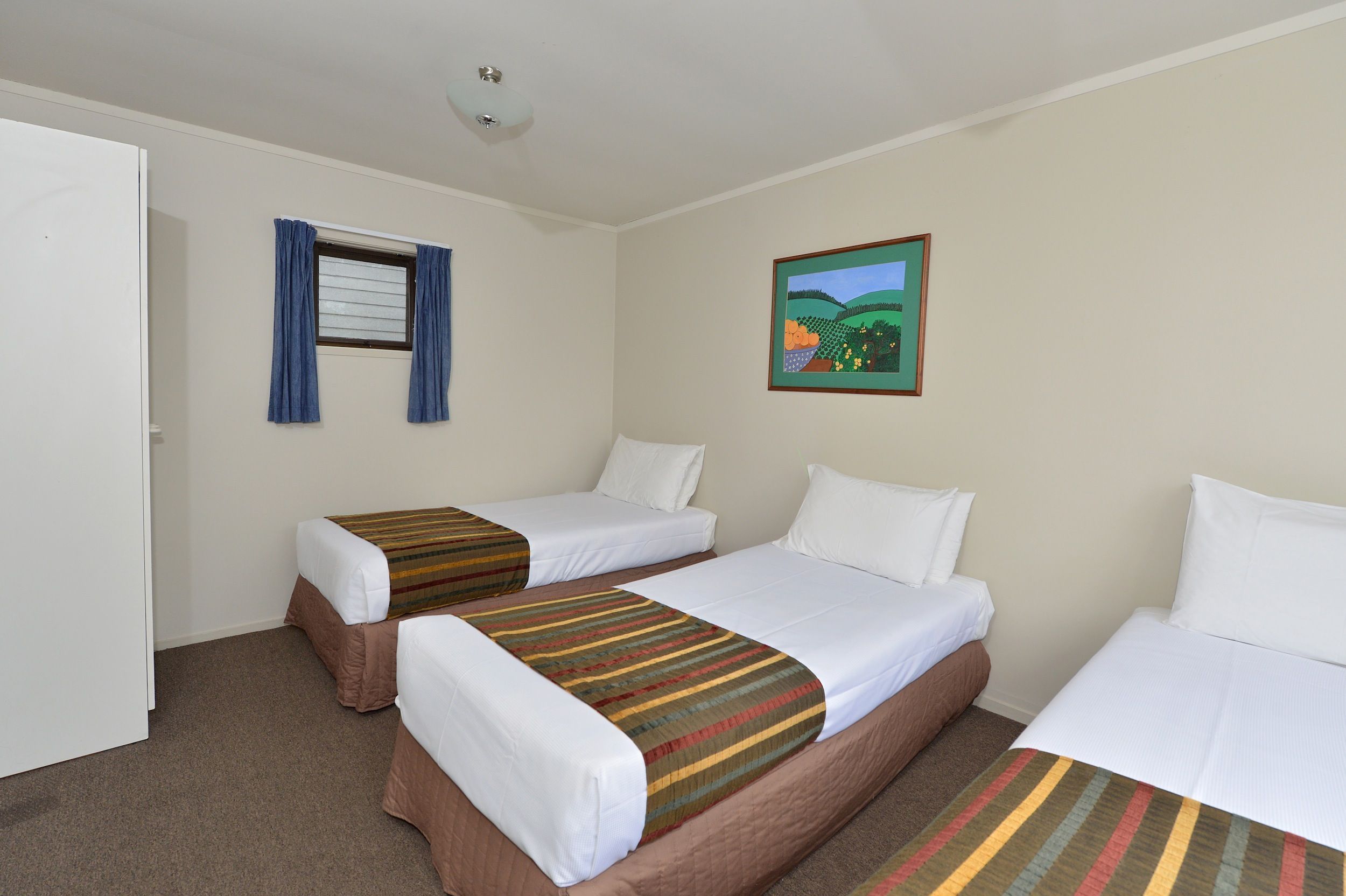 Quarto de hotel com três camas, uma janela com cortinas azuis, um quadro de paisagem e uma porta.