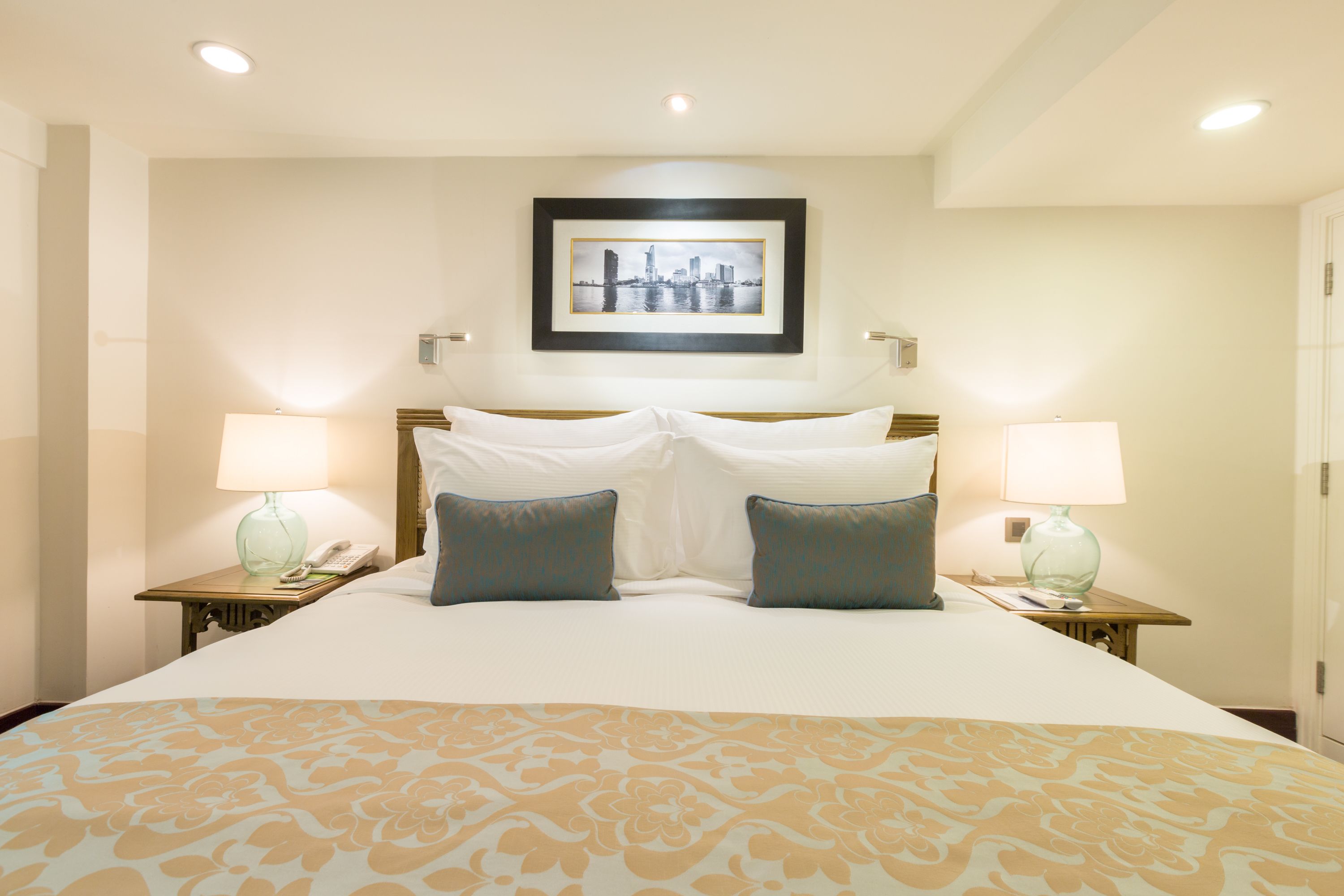 Quarto de hotel com uma cama grande, duas luminárias de cabeceira, uma imagem emoldurada da paisagem urbana na parede e um edredom estampado.