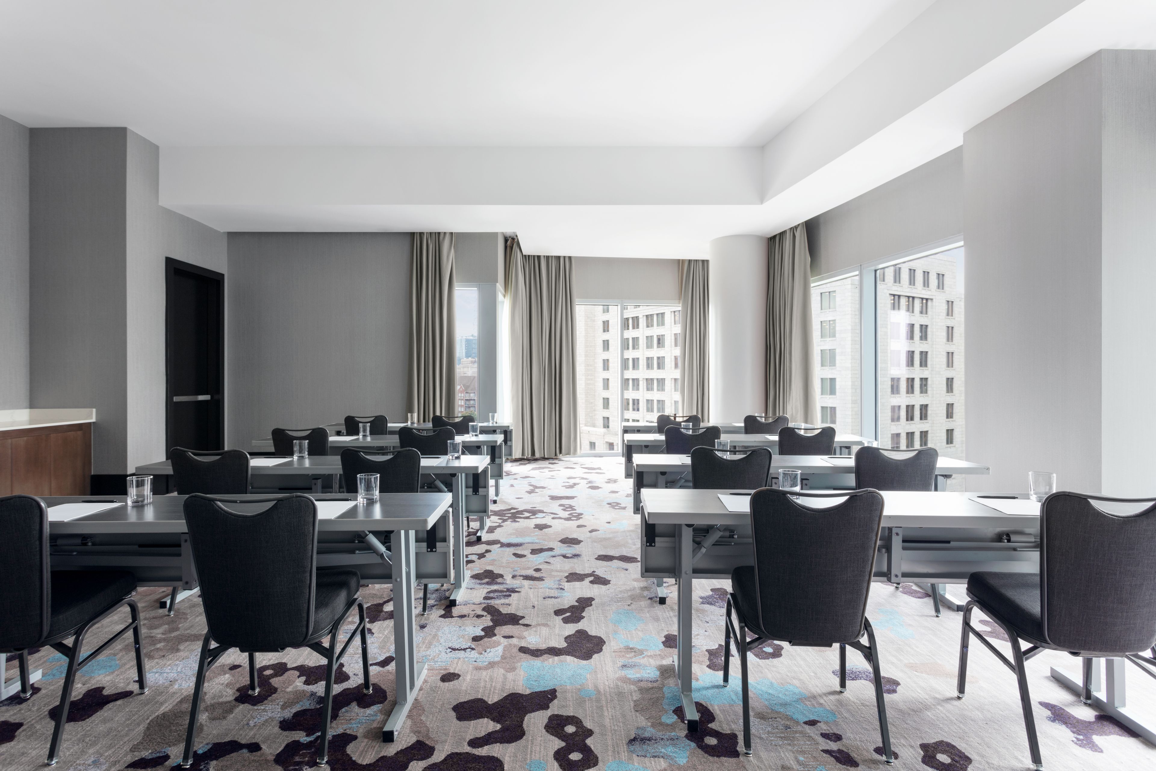 Uma sala de conferências de hotel com várias mesas e cadeiras, grandes janelas e um carpete estampado.