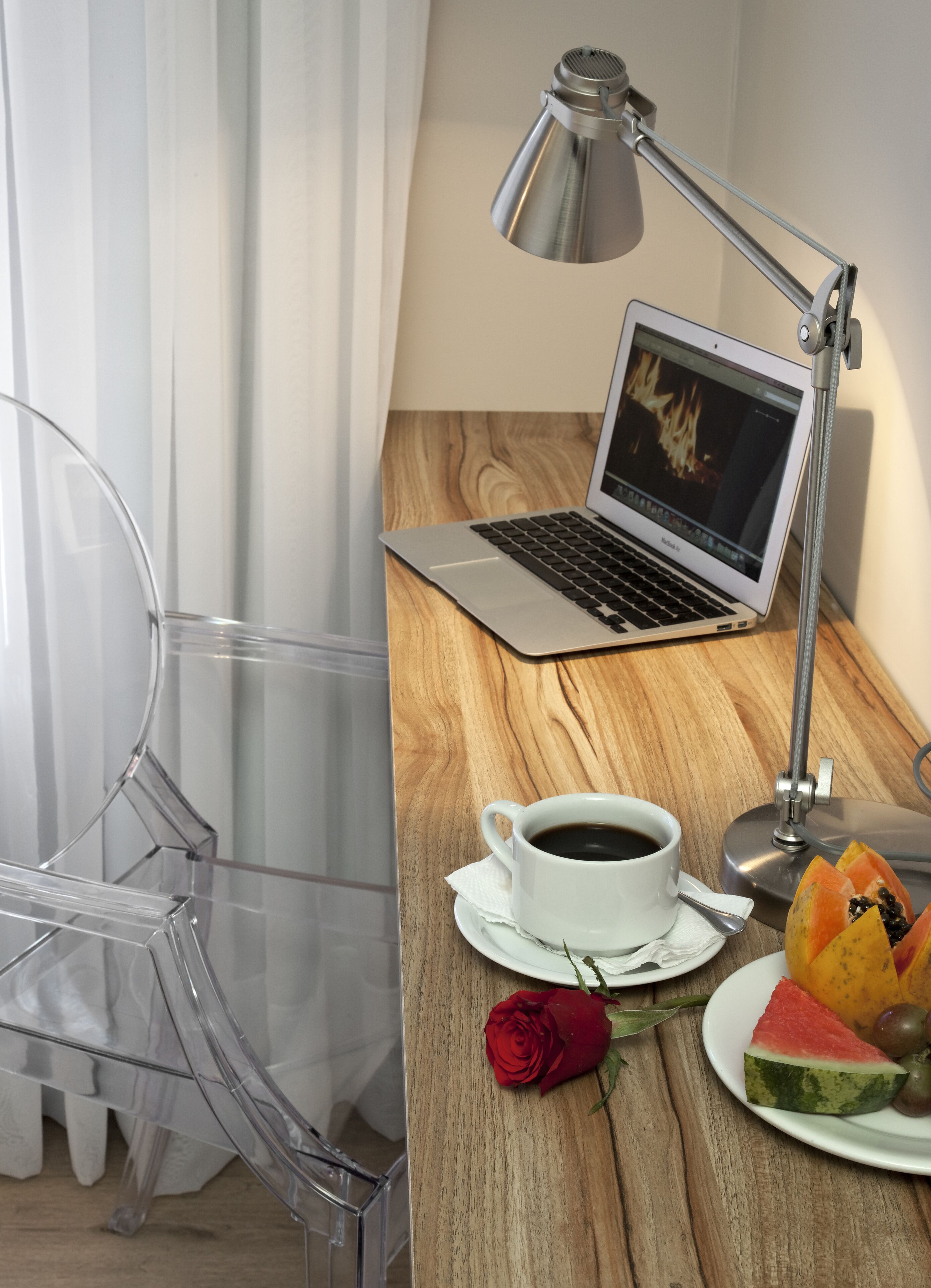 Uma mesa de madeira com um laptop, uma chávena de café, uma rosa e fruta.
