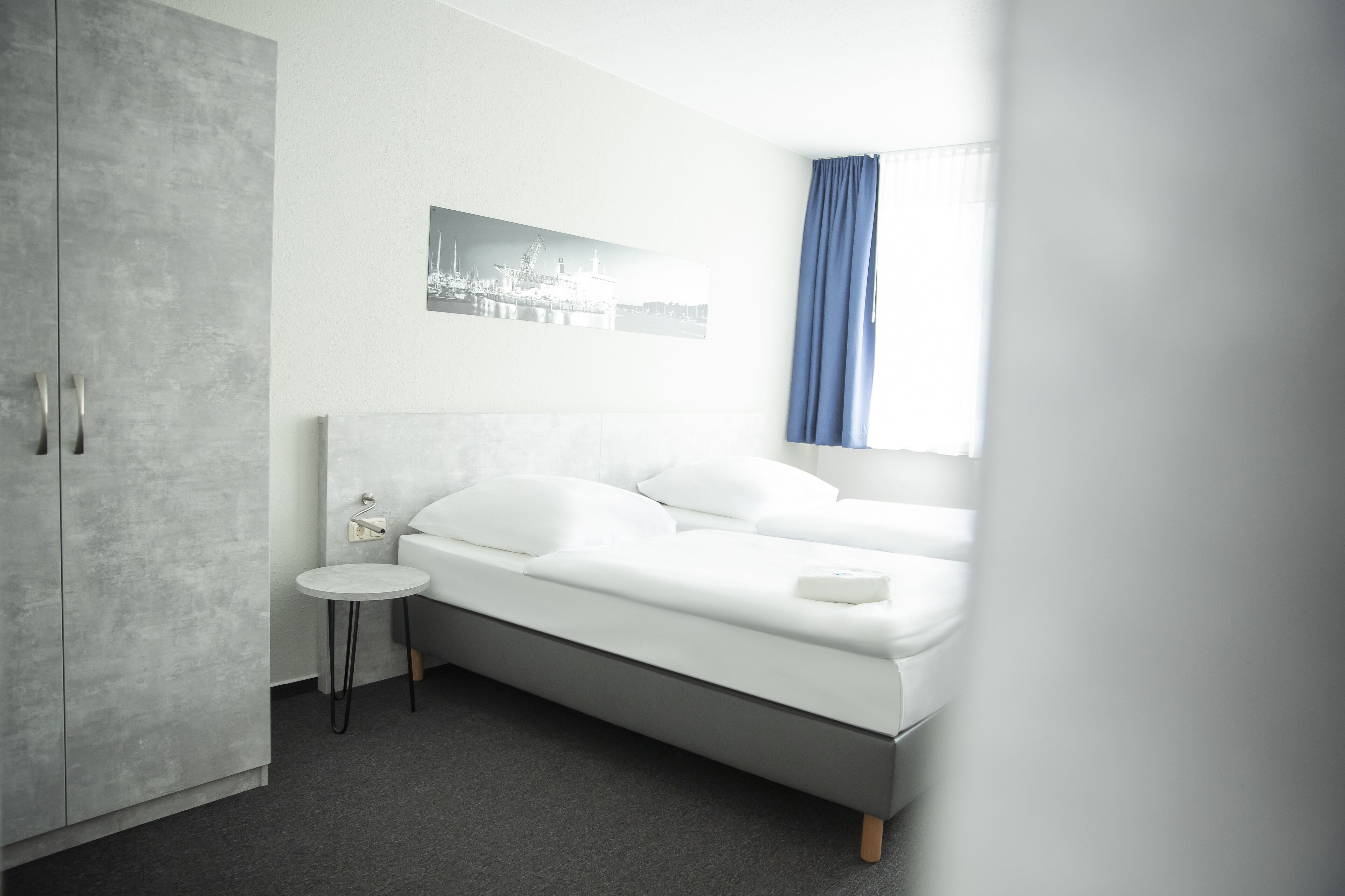 Quarto de hotel moderno com cama, mesa de cabeceira pequena, guarda-roupa e um quadro na parede.