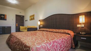 Quarto de hotel com uma cama grande, mesa de cabeceira com abajur, criado-mudo e televisão presa na parede.