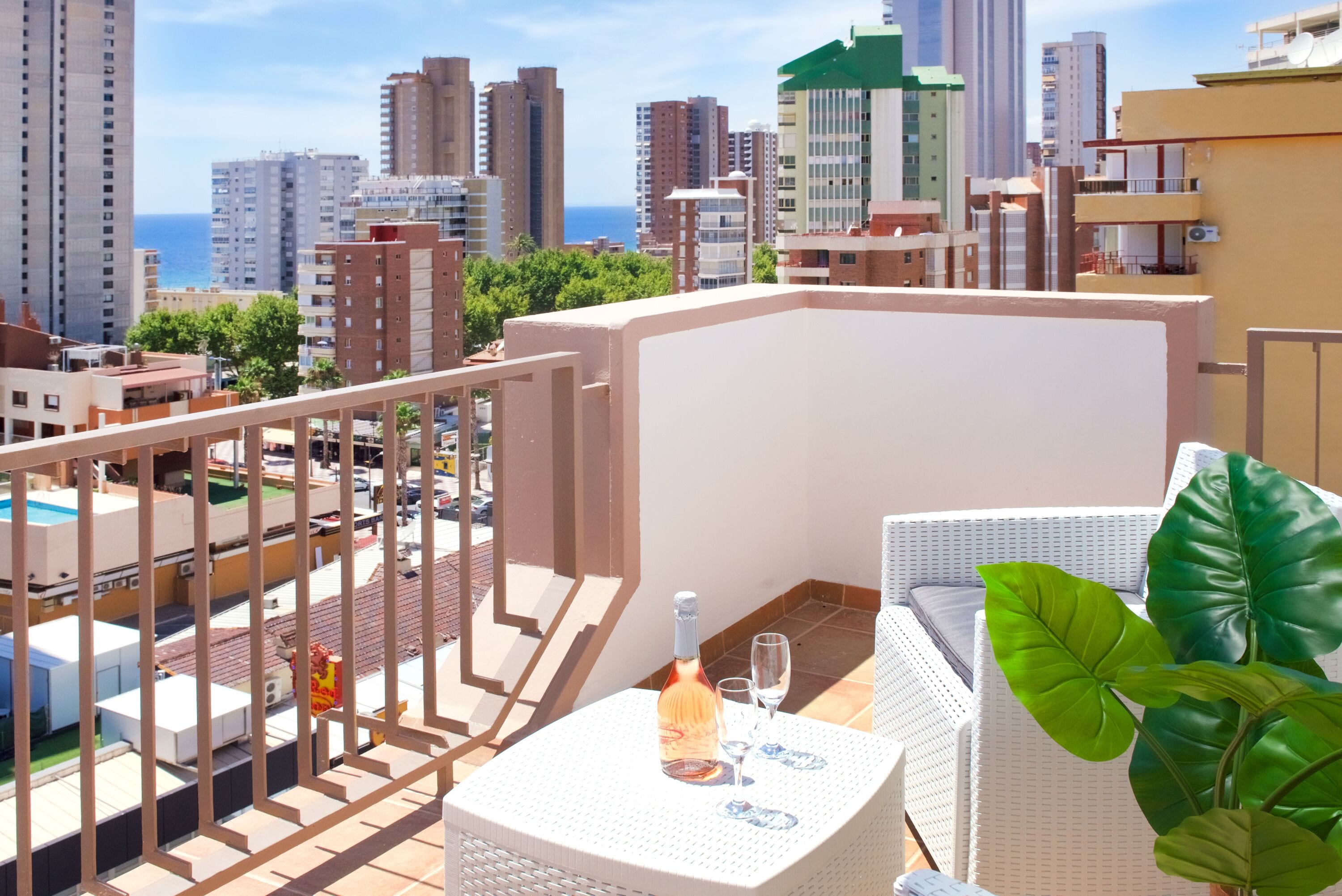 Varanda com sofá de vime branco, uma mesa pequena com garrafa e copos, e vista para a cidade e o oceano.