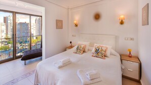 Quarto de hotel moderno com uma cama grande, duas luminárias de cabeceira, uma mesinha de cabeceira e vista para a paisagem urbana através de janelas do chão ao teto.