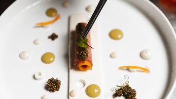 Um prato branco com um rolo de carne, guarnecido com caviar e uma folha verde, rodeado por pequenos pontos brancos e amarelos.