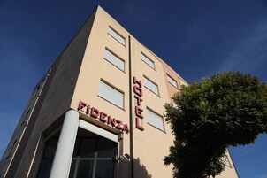 Um edifício de hotel com fachada bege, uma placa grande com a inscrição 'FIDENZA HOTEL' e uma árvore em primeiro plano.