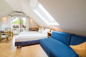 Quarto com claraboia, cama, sofá azul e vista para uma varanda florida.