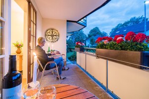 Um homem sentado à mesa de madeira numa varanda, com uma vela, uma garrafa de vinho e copos. Há plantas e flores em vasos na varanda.