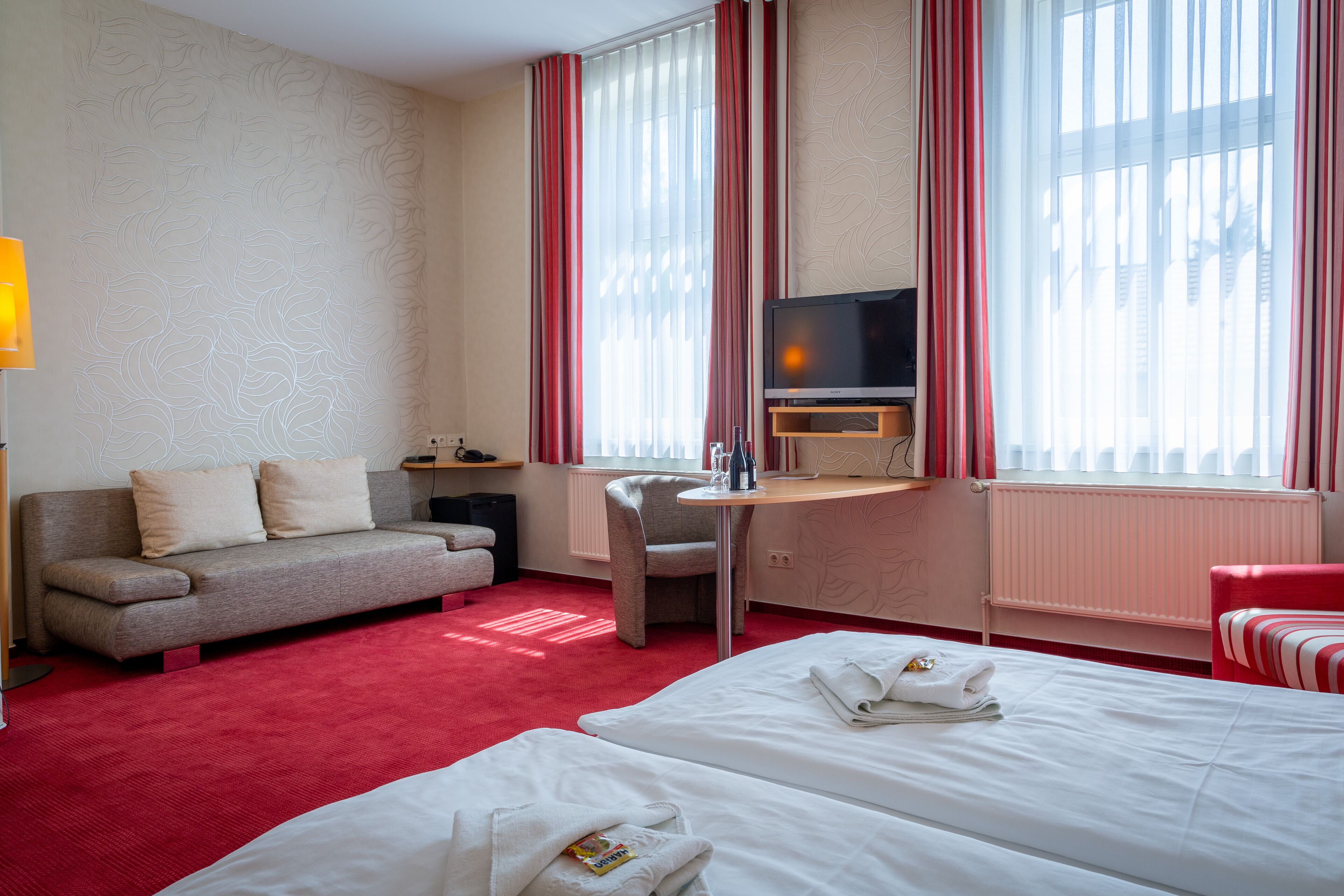 Quarto de hotel com carpete vermelho, sofá, escrivaninha com televisão, cadeira e duas camas.
