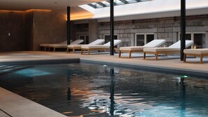 Uma área interna de piscina moderna, com espreguiçadeiras, teto de vidro transparente e parede de pedra ao fundo.