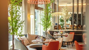 Restaurante com decoração moderna, apresentando uma variedade de assentos, incluindo um banco curvo e cadeiras individuais. Vasos com plantas adicionam verde ao ambiente.