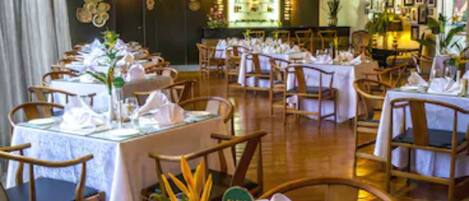 Um restaurante com várias mesas preparadas para refeições, com toalhas de mesa brancas e louças bem organizadas.