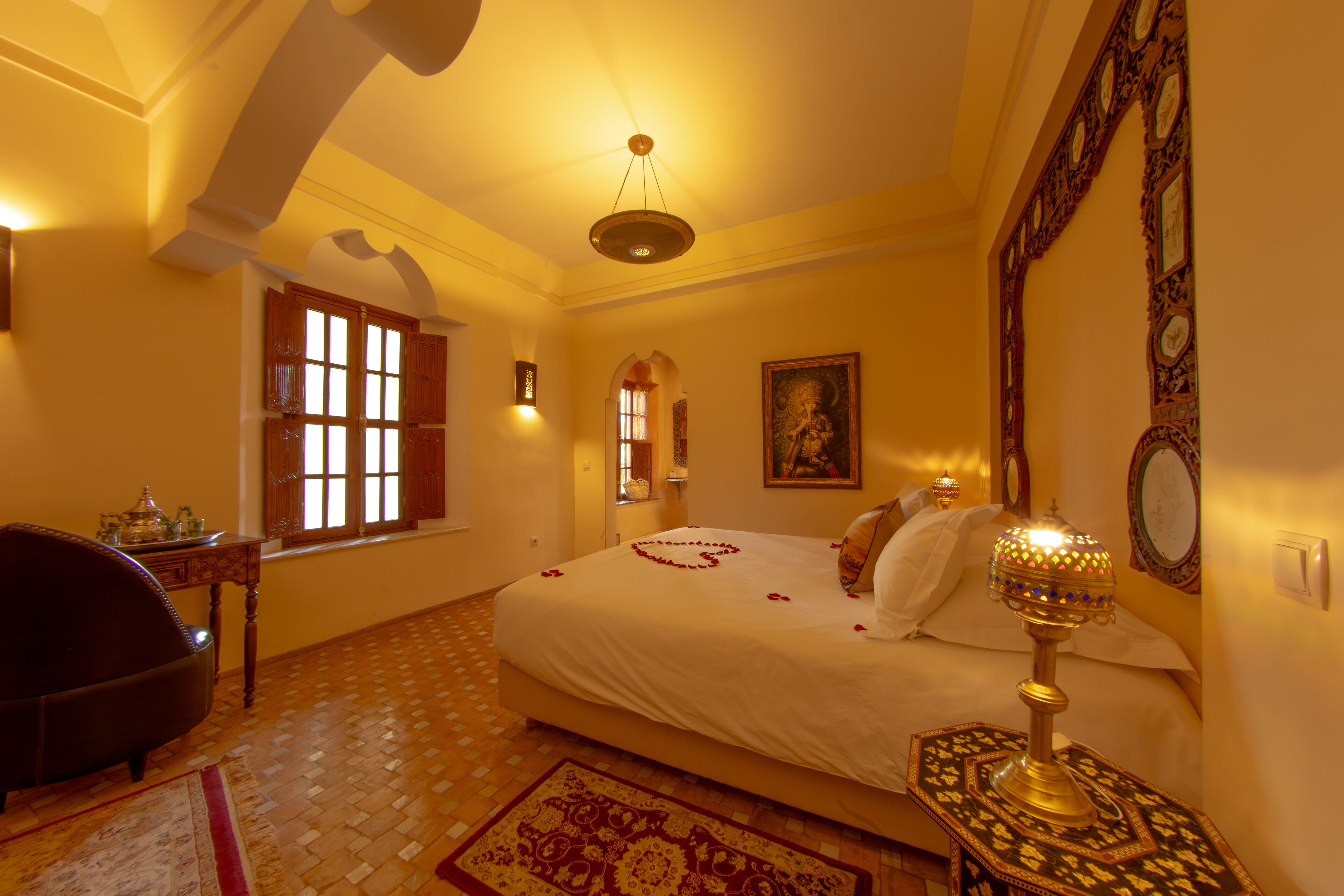 Quarto de hotel com cama, mesa de cabeceira, cadeira, janela com persianas e um objeto decorativo na parede.