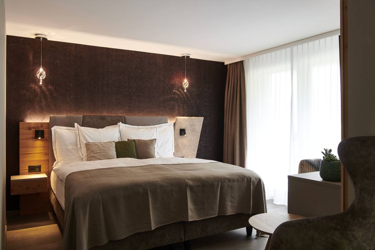 Quarto de hotel moderno com uma cama grande, mesas de cabeceira, uma mesinha lateral e um vaso de plantas.