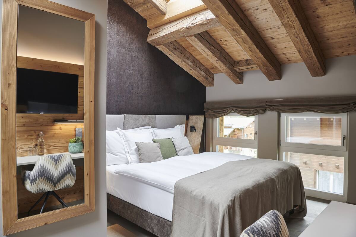 Quarto com cama grande, teto de madeira, escrivaninha com cadeira e janela com cortinas.