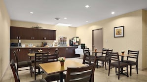 Uma sala de jantar de hotel com uma mesa central rodeada por cadeiras, área de cozinha com frigorífico e máquina de café, e um quadro na parede.