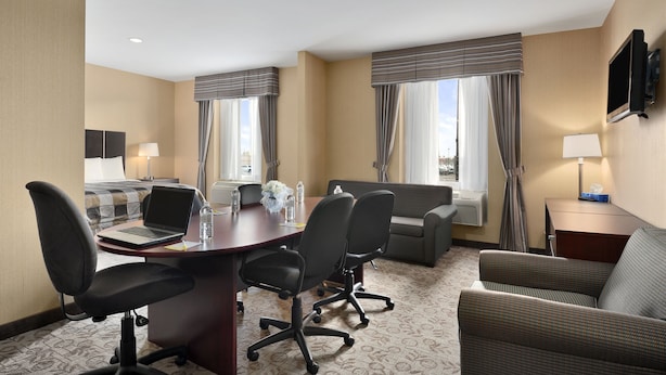 Quarto de hotel com mesa de conferência, duas cadeiras de escritório, sofá, televisão e janela com cortinas.
