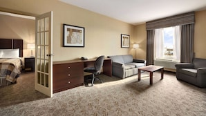 Quarto de hotel com cama, escrivaninha, sofá, poltrona, mesa de centro e janela com cortinas.
