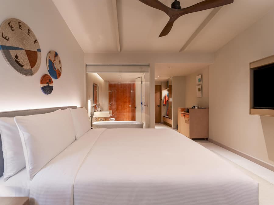 Quarto de hotel com uma cama grande, ventoinha de madeira no teto, banheiro com chuveiro e TV fixada na parede.