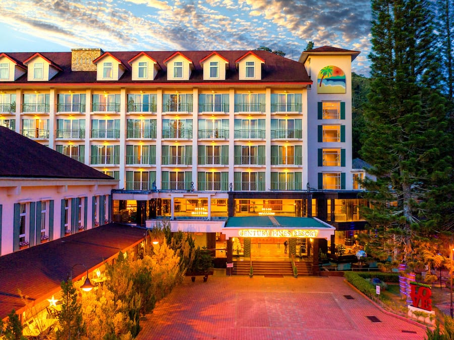 Um hotel com vários andares, entrada central, varandas e uma placa com a inscrição "CENTURY PINES RESORT".