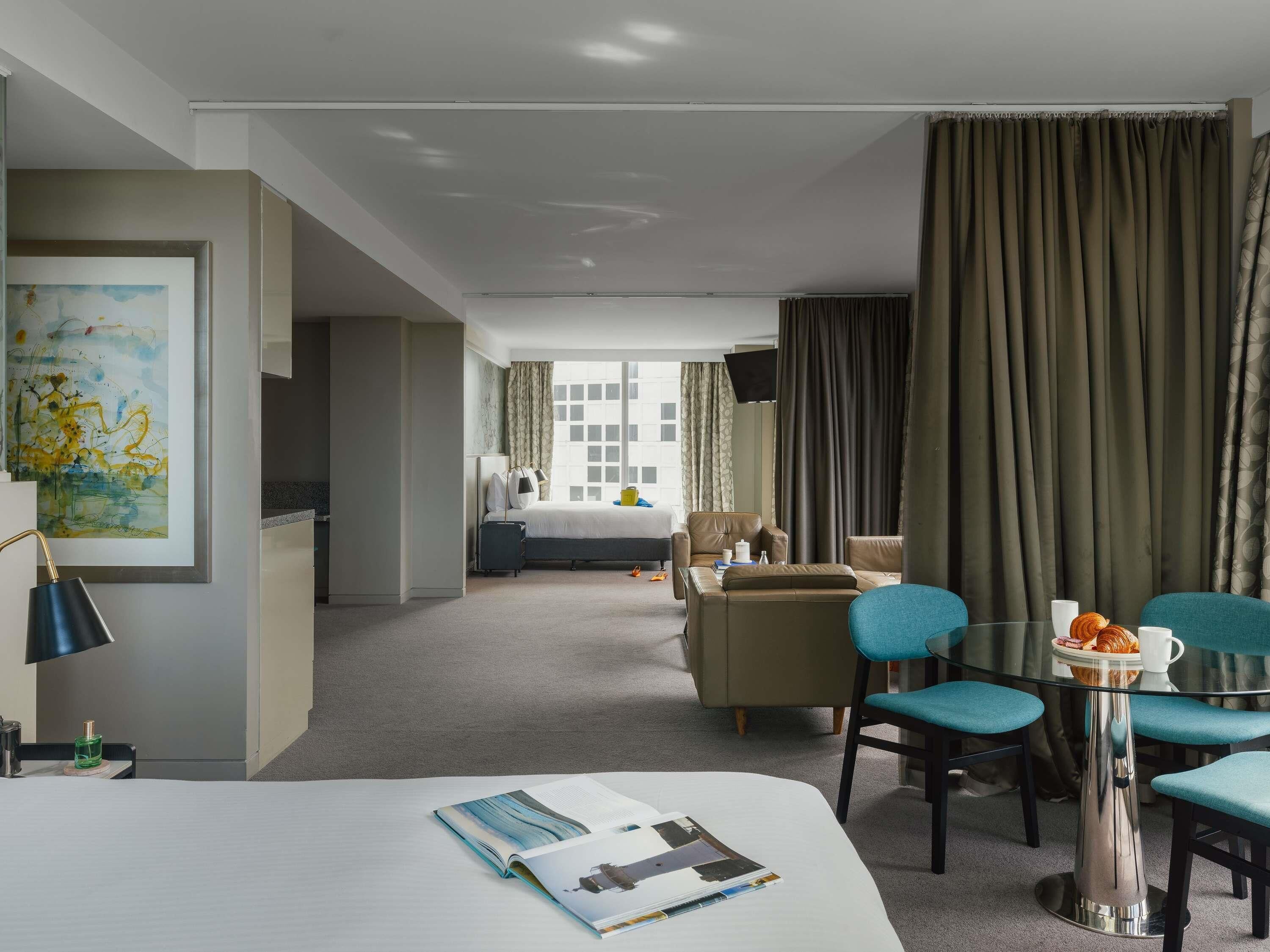 Quarto de hotel com uma cama, uma escrivaninha com abajur, uma cadeira, uma mesa com uma xícara e vista para outro quarto.