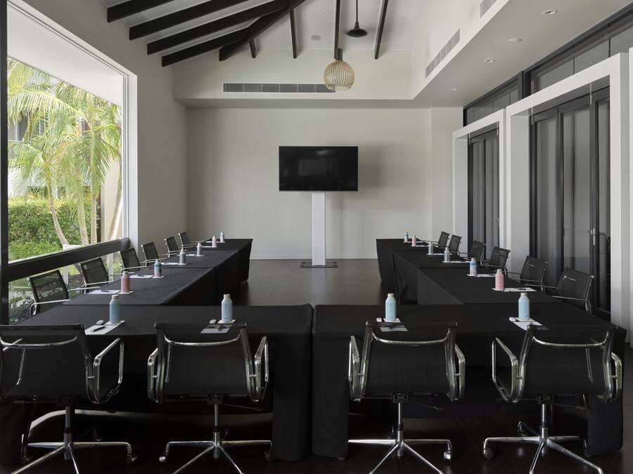 Sala de conferências com uma mesa comprida, cadeiras, tela de TV e janelas amplas com vista para área verde.