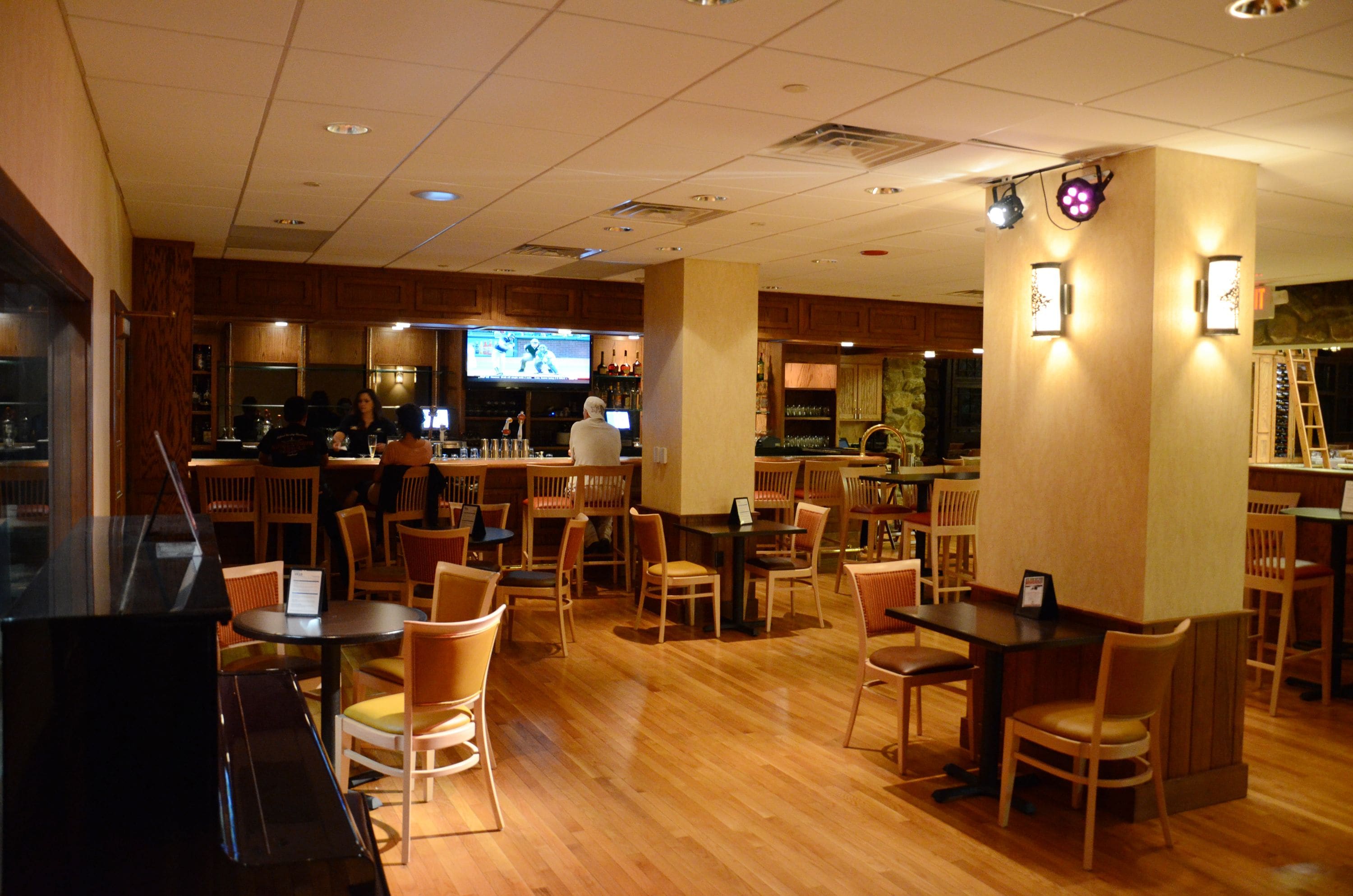 Restaurante com piso de madeira, várias mesas e cadeiras, um balcão e uma televisão exibindo um evento esportivo.