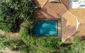Vista aérea de uma piscina retangular rodeada por um deck de madeira, com vegetação densa e um muro de tijolos ao fundo.
