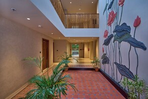 Corredor de hotel com chão em azulejo, plantas em vasos, corrimão de madeira e um mural na parede.