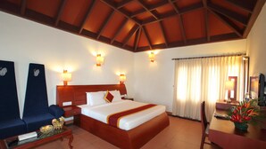 Quarto de hotel com uma cama grande, cabeceira de madeira, duas luminárias de apoio, uma mesa com cadeira e uma pequena mesa com abajur, além de uma janela com cortinas.