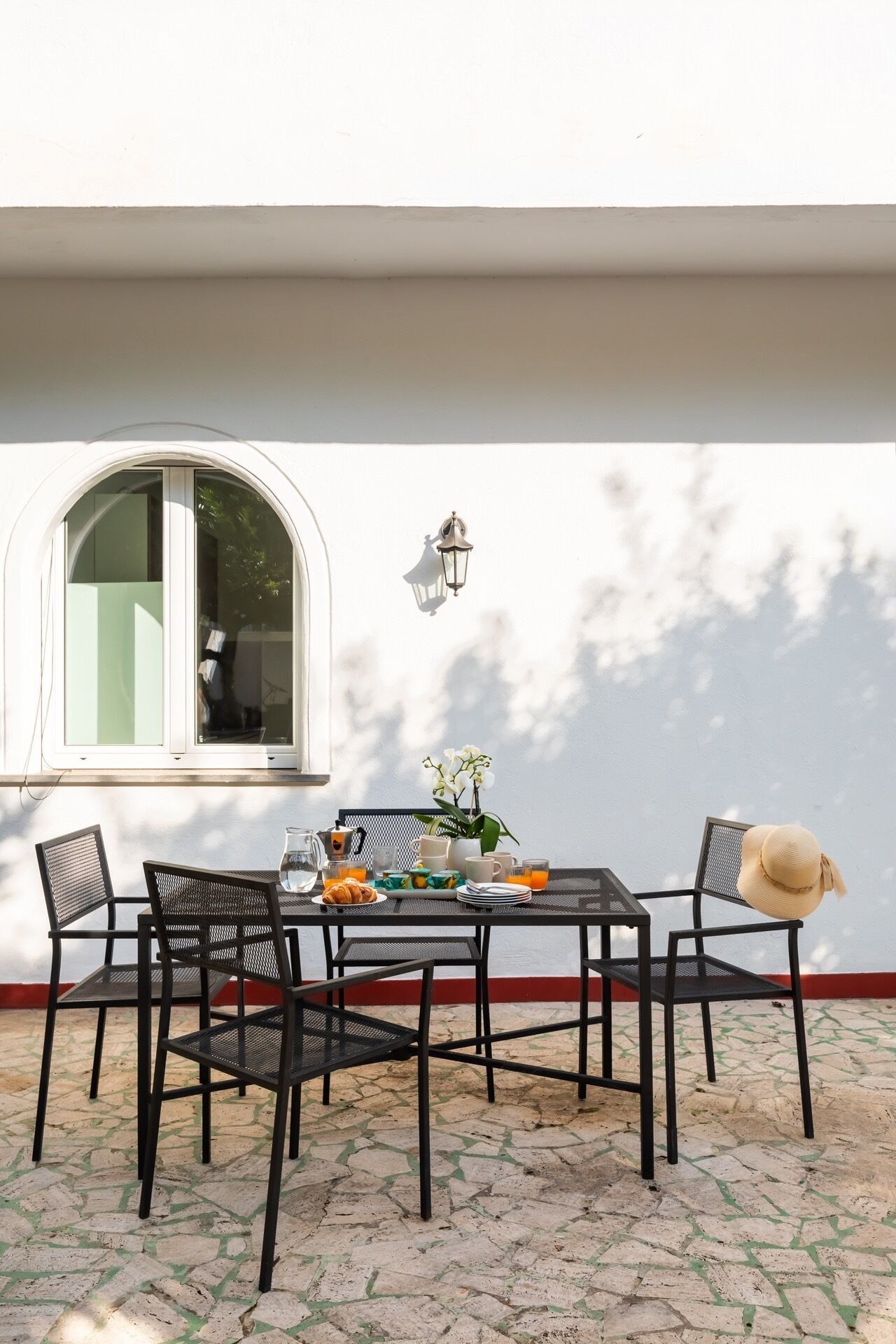 Uma área de refeições ao ar livre com mesa, cadeiras, uma janela e um aparelho de luz fixado na parede.