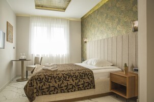 Quarto de hotel com cama, mesa de cabeceira, cadeira, pequena mesa com abajur e janela com cortinas leves.