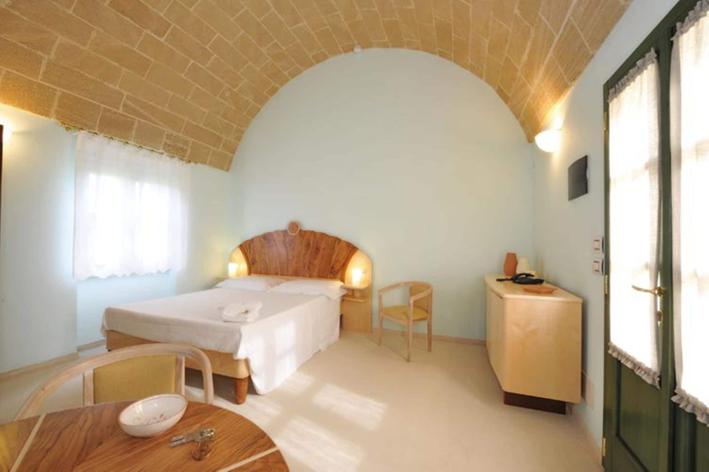 Quarto com cabeceira de madeira, uma cama de solteiro, mesa de cabeceira, uma cadeira, um pequeno armário e janela com cortinas.