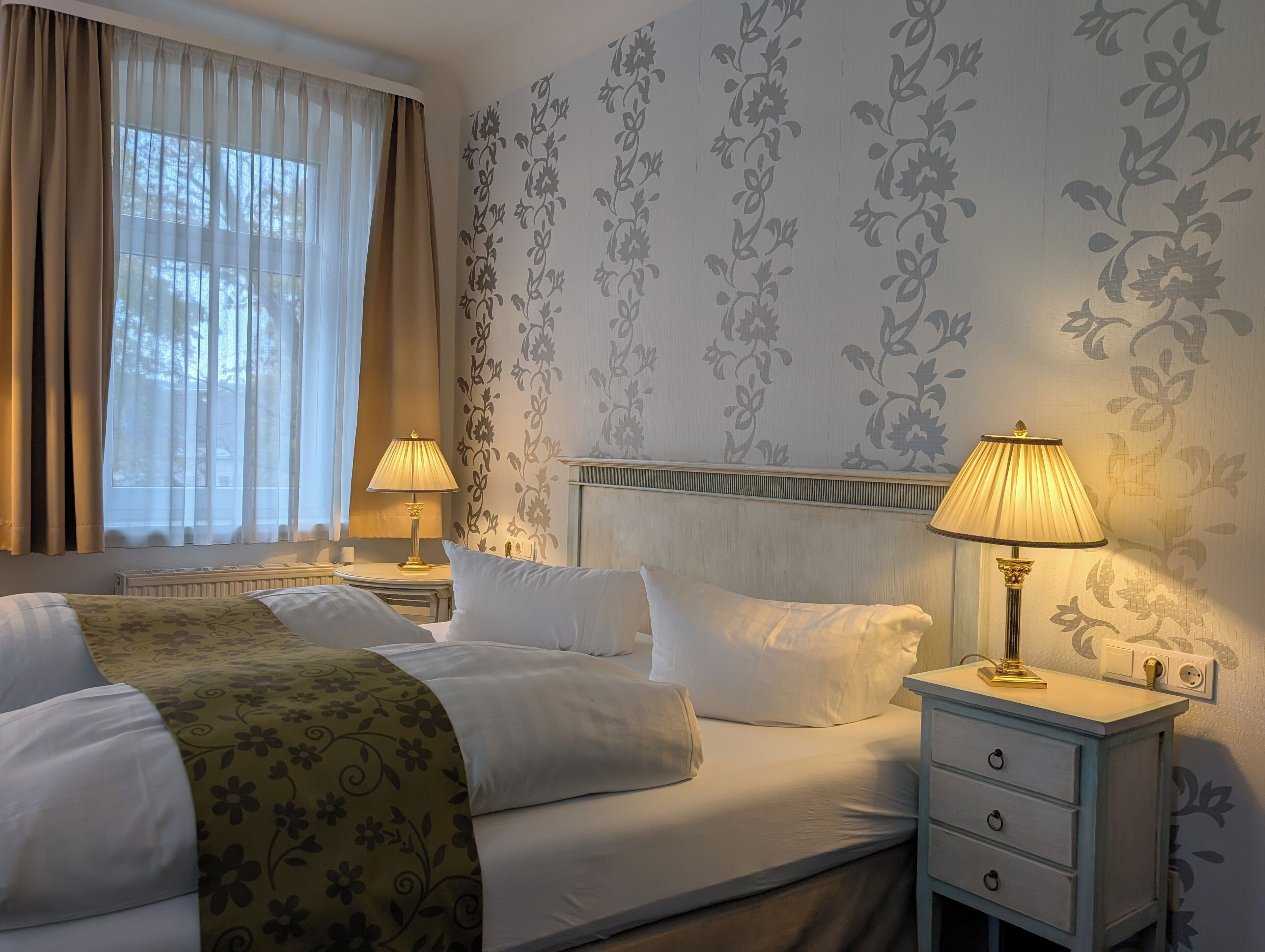 Quarto de hotel com uma cama grande, duas mesas de cabeceira com abajures, papel de parede floral e uma janela com cortinas leves.