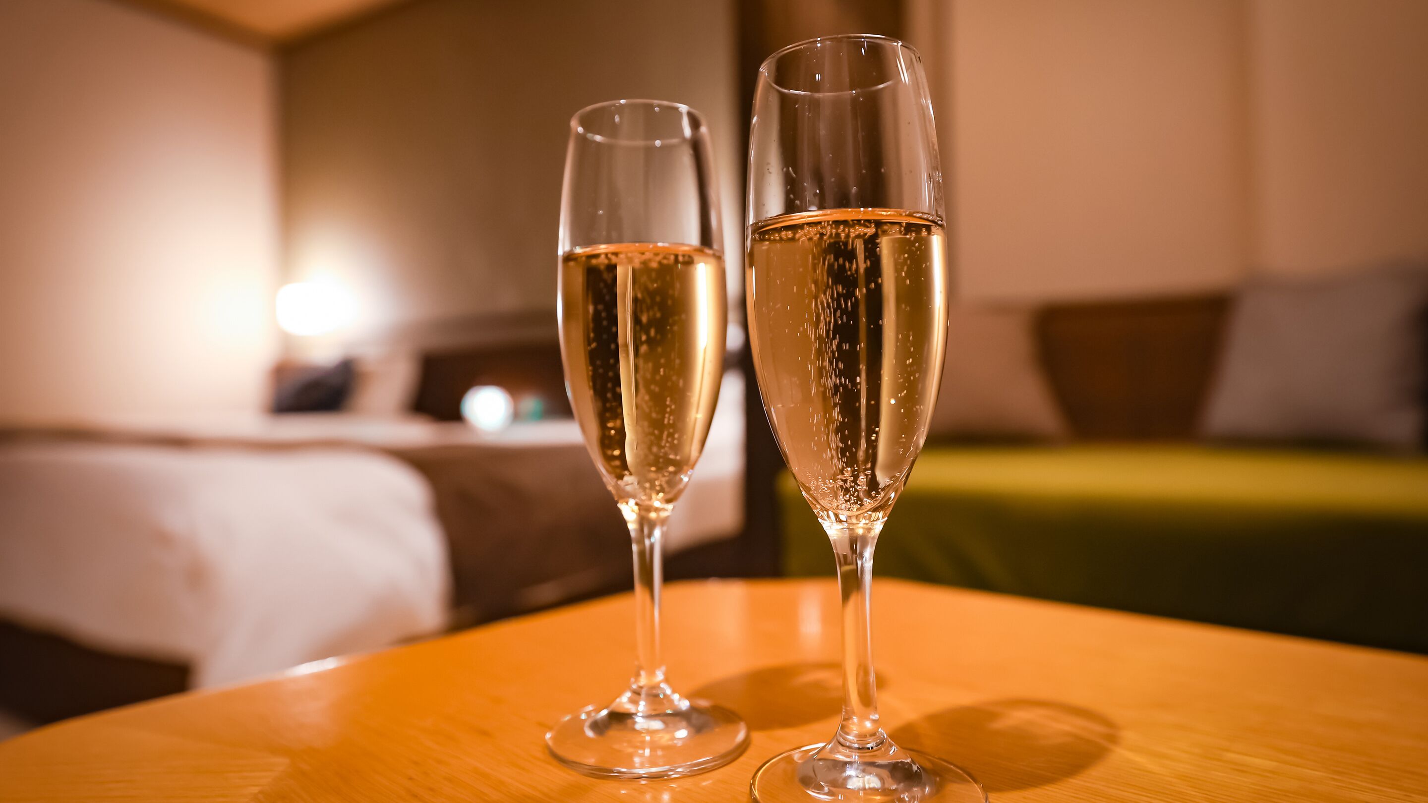 Duas taças de champanhe sobre uma mesa de madeira em quarto de hotel, com uma cama e mesa de cabeceira ao fundo.