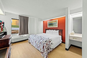 Quarto de hotel com cama, escrivaninha, televisão, janela com cortinas, um quadro na parede e espelho no banheiro.
