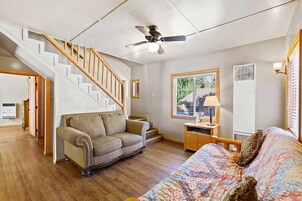 Uma sala de estar ampla com escada de madeira, sofá bege, cama colorida e ventilador de teto.