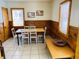 Uma zona de refeições com mesa e cadeiras de madeira, uma cesta de vime e uma janela com persianas.