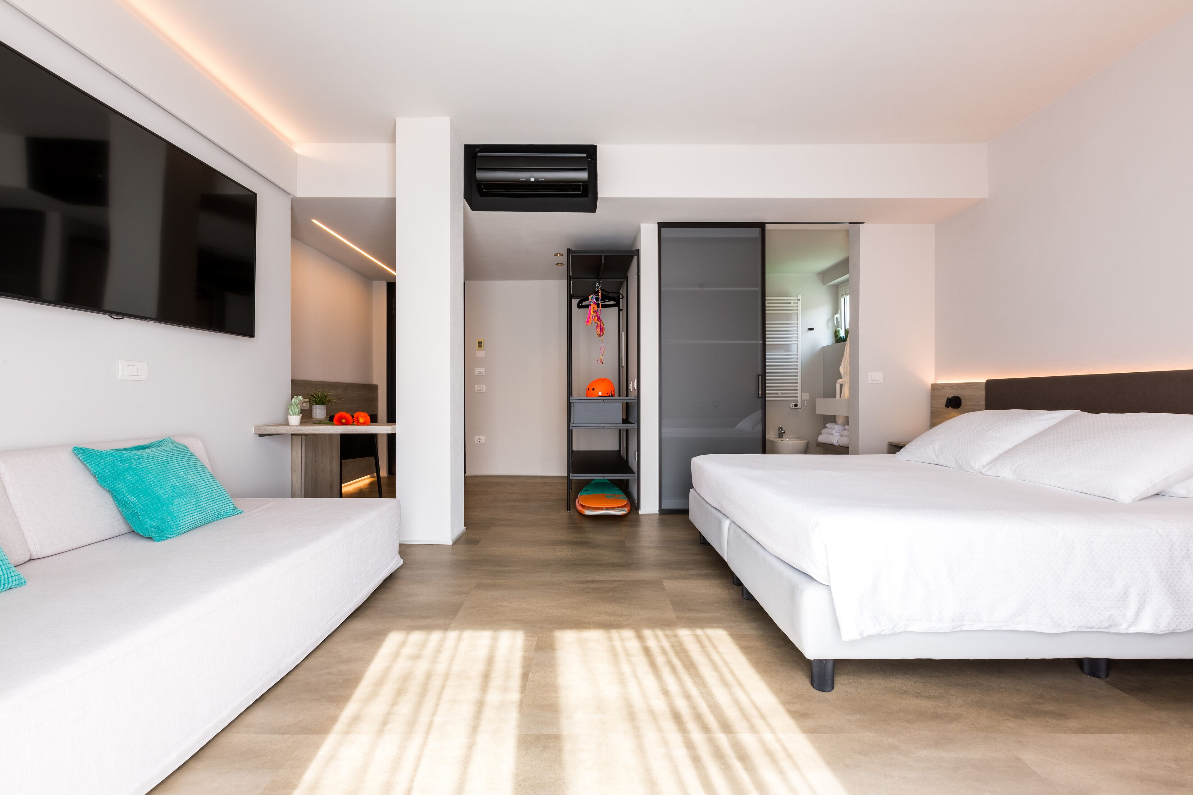 Quarto de hotel moderno com sofá, cama, televisão, guarda-roupa e uma pequena área de cozinha.