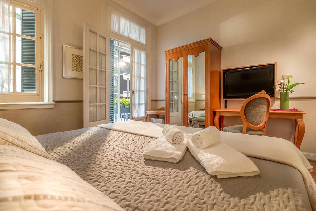 Quarto de hotel com cama, escrivaninha com televisão, cadeira e janela com persianas.