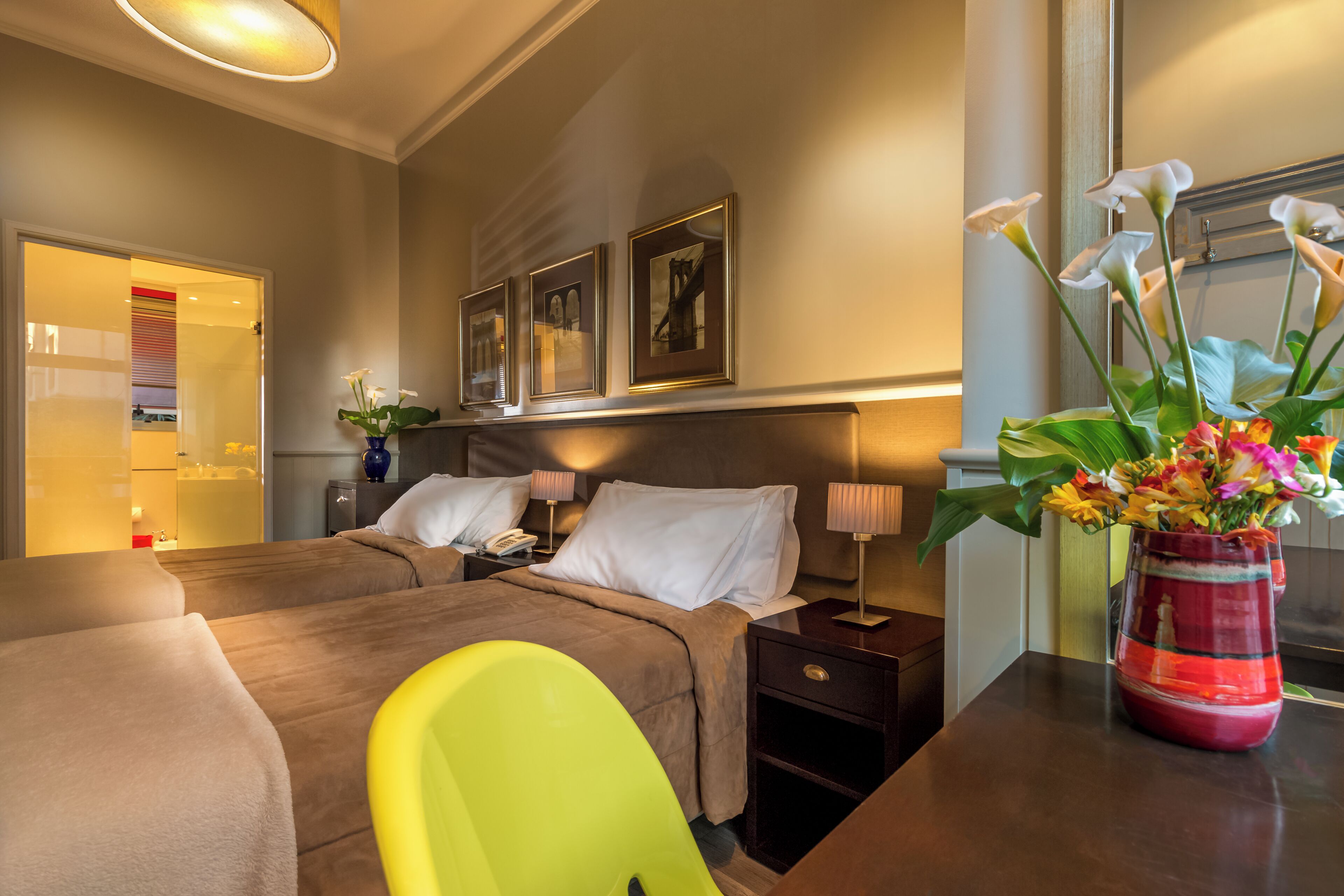 Quarto de hotel com cama, mesas de cabeceira, escrivaninha, cadeira e um vaso com flores.