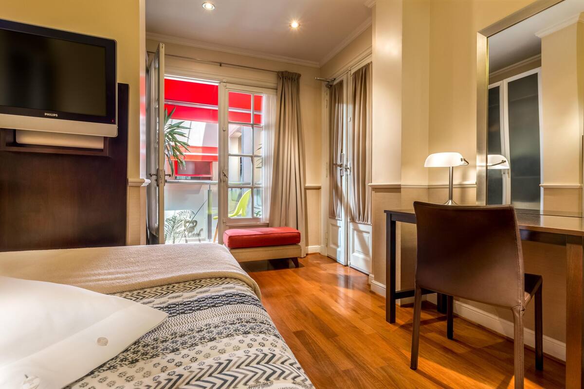 Quarto de hotel com uma cama, uma escrivaninha com cadeira, televisão e janela com cortinas.