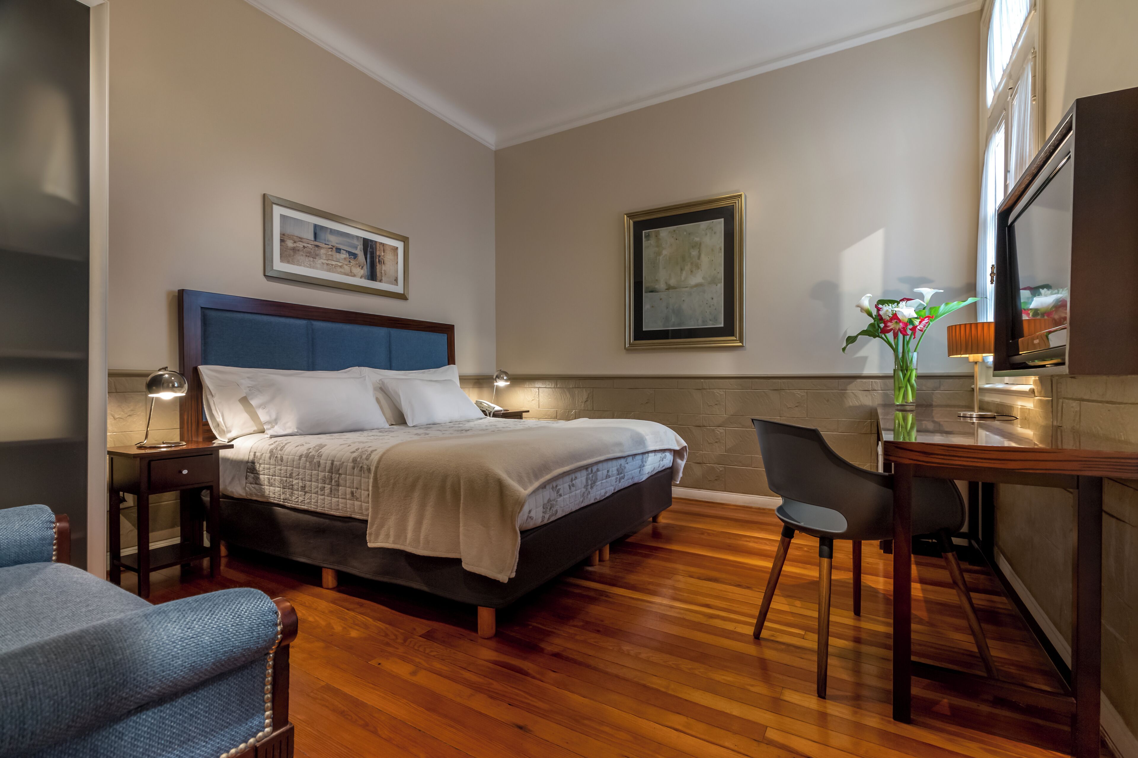 Quarto com cama, mesas de cabeceira, escrivaninha e uma cadeira, além de uma janela.