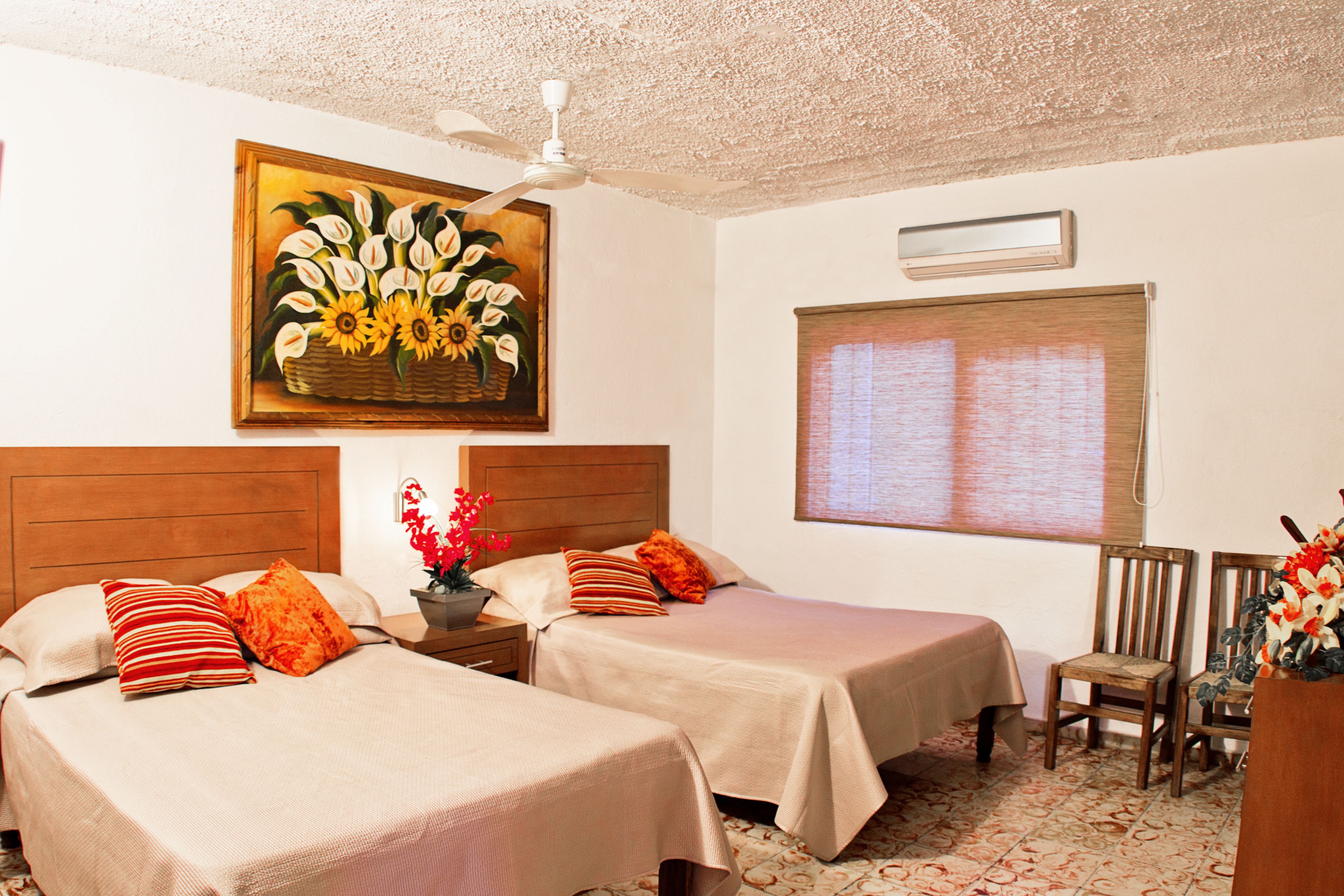 Quarto de hotel com duas camas, um ventilador de teto, um quadro de flores, uma janela com persianas e uma cadeira de madeira.
